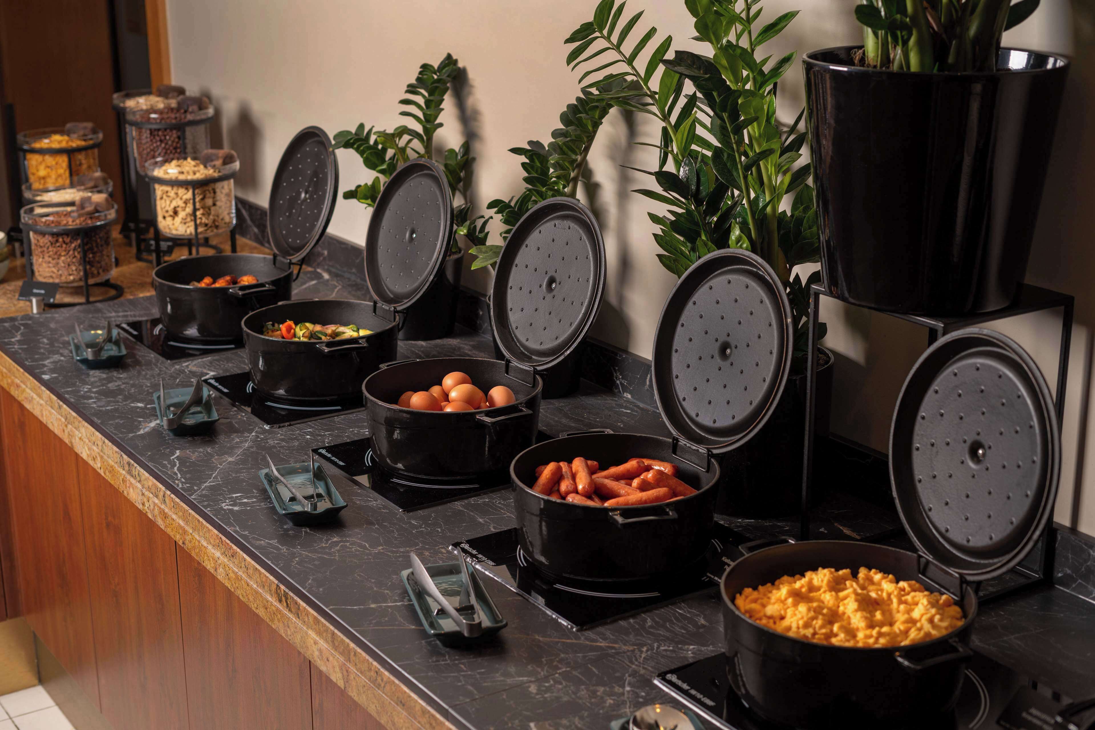 Buffet montado com diversos alimentos em panelas pretas e uma planta em uma panela preta.
