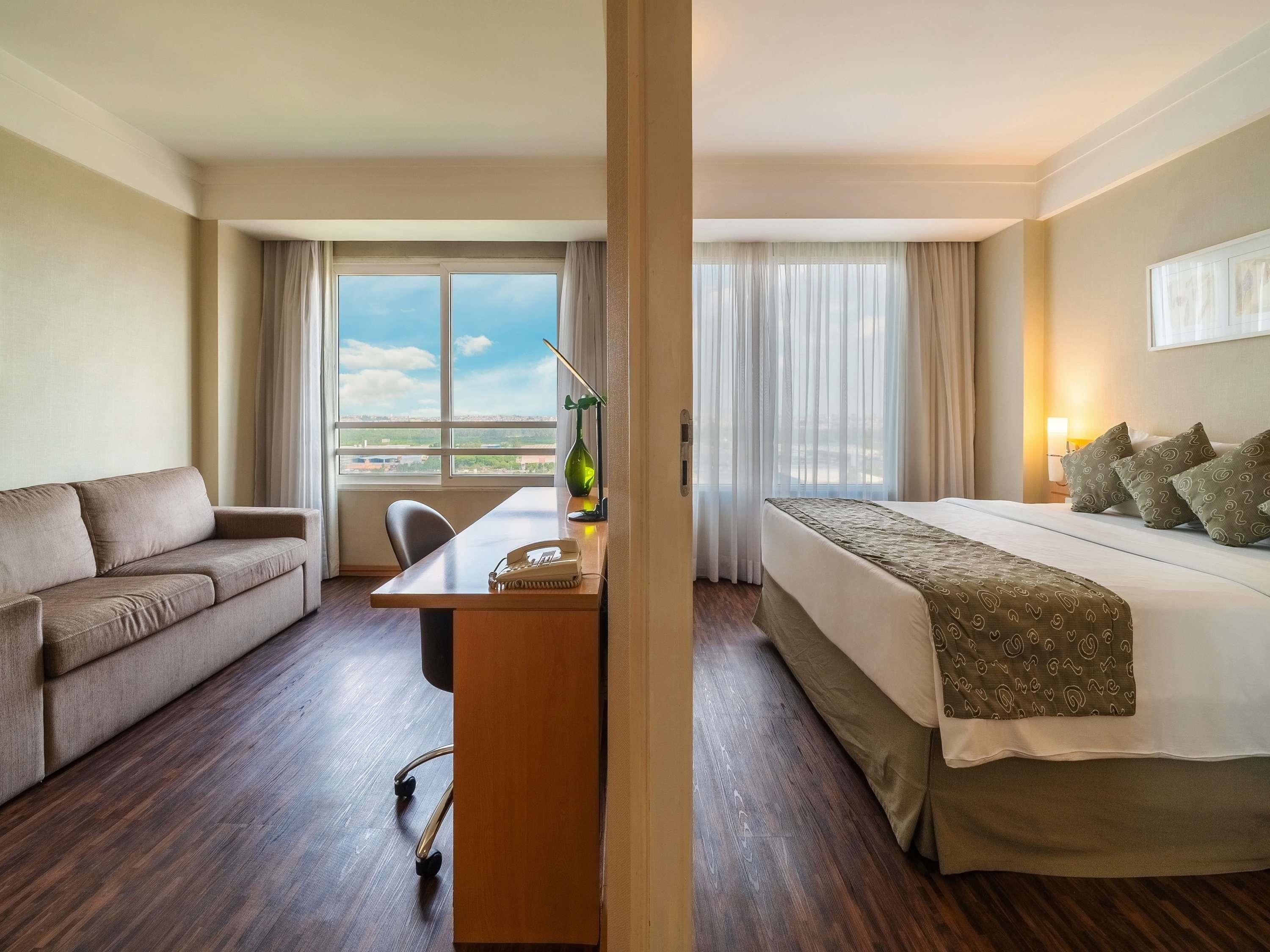 Quarto de hotel com cama, sofá, escrivaninha e janela com cortinas.