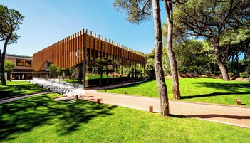 Um edifício moderno com fachada de madeira e entrada de vidro, rodeado por um gramado bem cuidado e árvores.