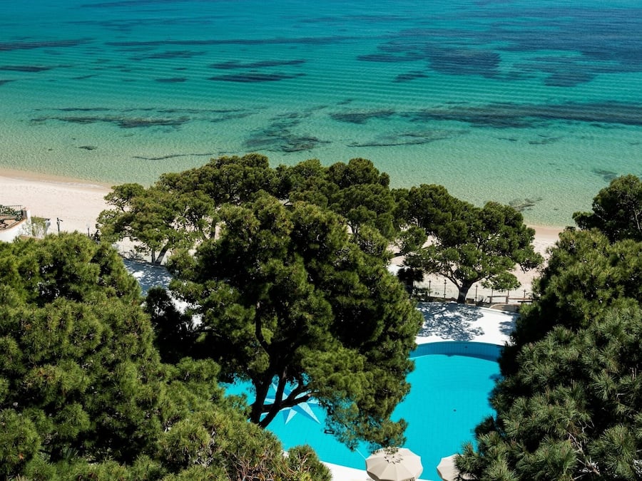 Vista aérea de um resort à beira-mar com piscina, rodeada por árvores, e vista para o mar azul e límpido.
