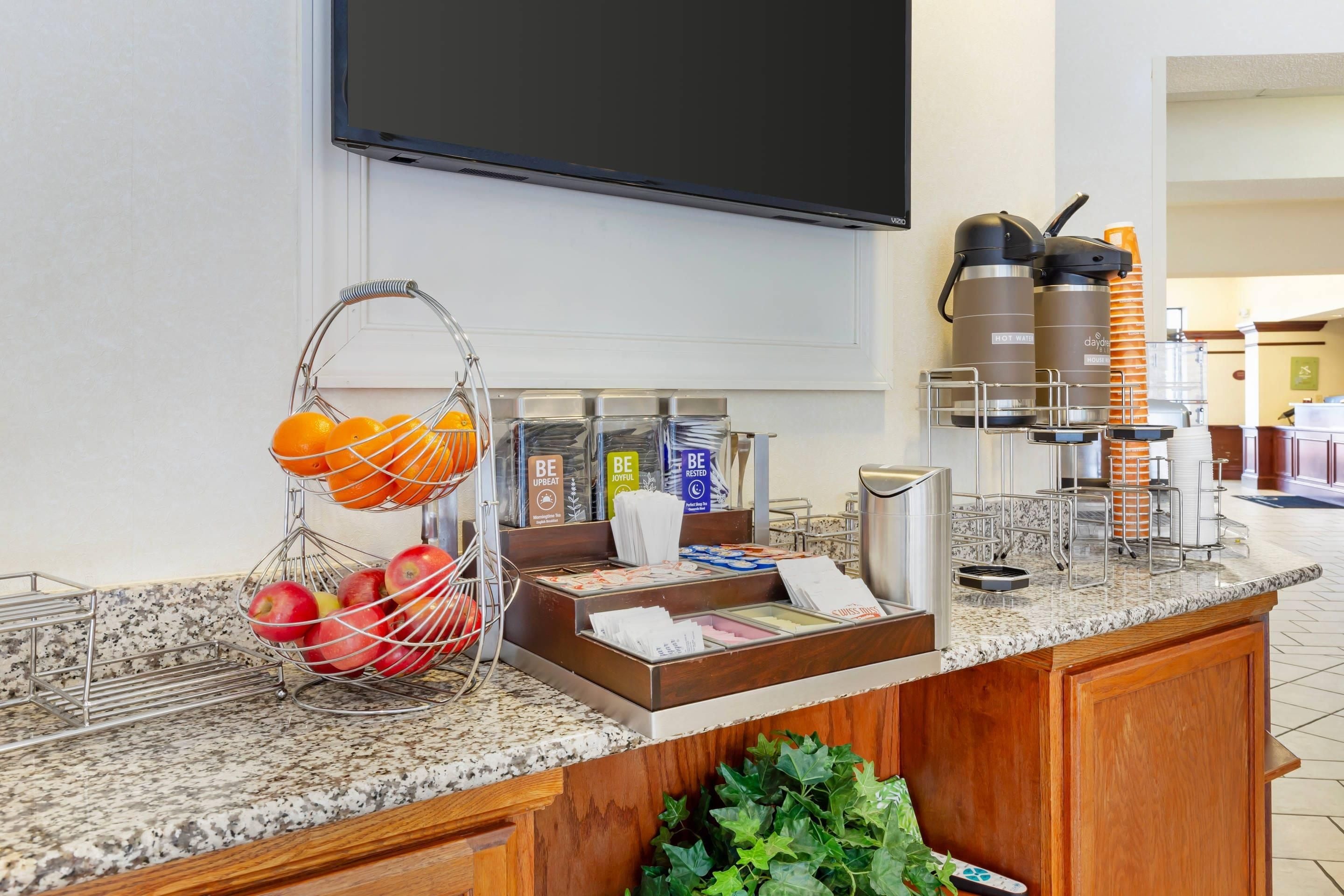 Buffet de café da manhã em hotel, com cestas de frutas, máquinas de café e uma TV de tela plana.