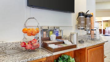 Buffet de café da manhã em hotel, com cestas de frutas, máquinas de café e uma TV de tela plana.