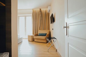 Uma sala de estar moderna com um sofá bege, uma mesinha de apoio, uma televisão fixada na parede e uma janela com cortinas.