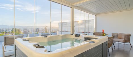 Área interna moderna com hidromassagem e vista panorâmica da cidade, grandes janelas e um espaço de estar com móveis de vime.
