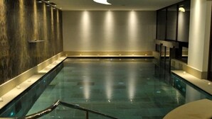 Uma área de piscina interior moderna, com barreira de vidro, degraus de pedra e um candeeiro de teto singular.