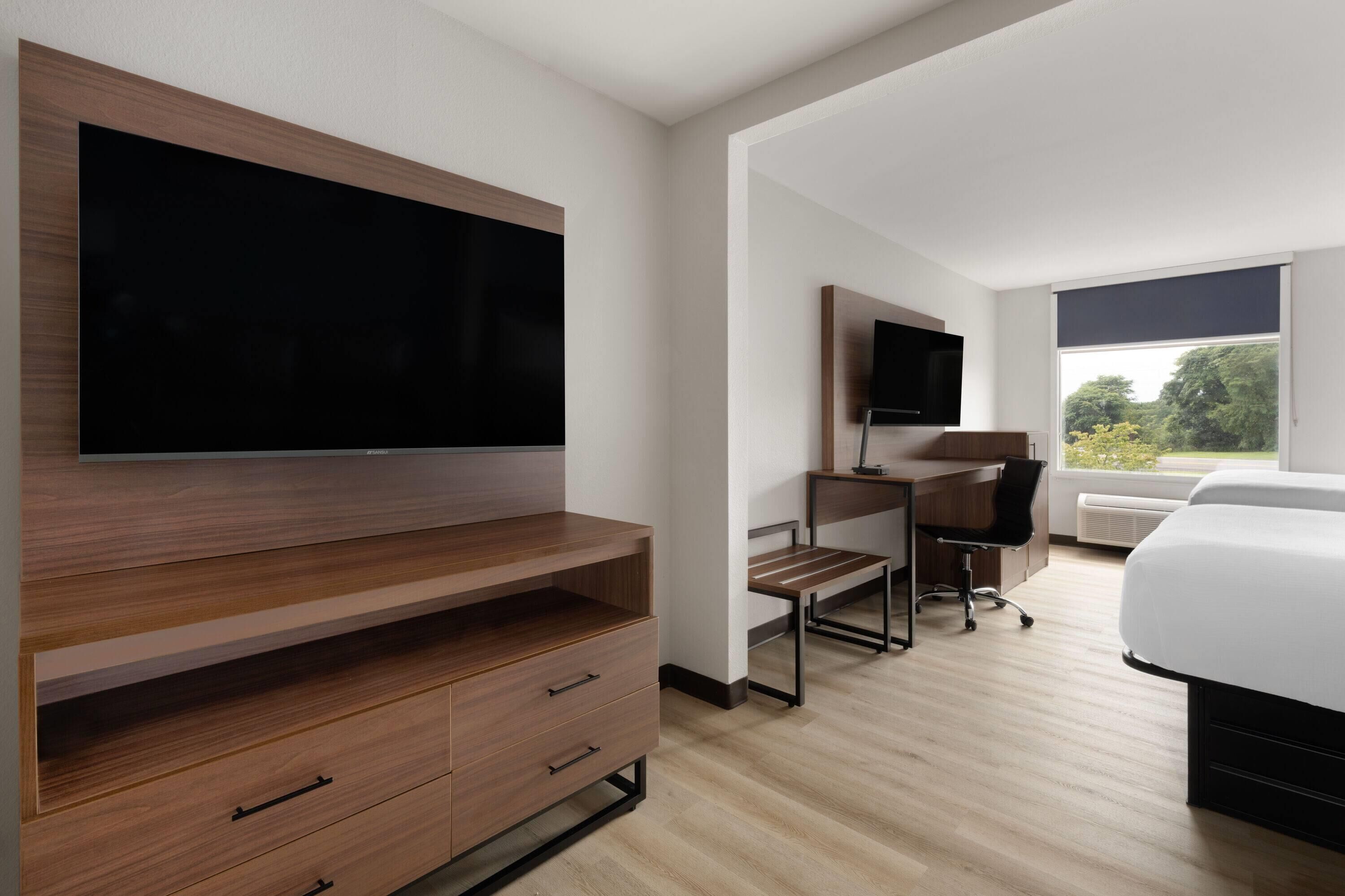Quarto de hotel com uma televisão de tela plana grande fixada em um móvel de madeira, uma escrivaninha com cadeira e uma cama com colchão branco.
