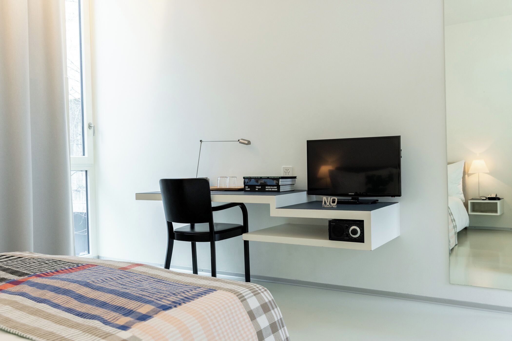 Quarto de hotel moderno com cama, escrivaninha com televisão, cadeira e mesa de cabeceira com abajur.
