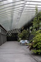 Uma galeria coberta com teto metálico ondulado, bancos e vegetação.
