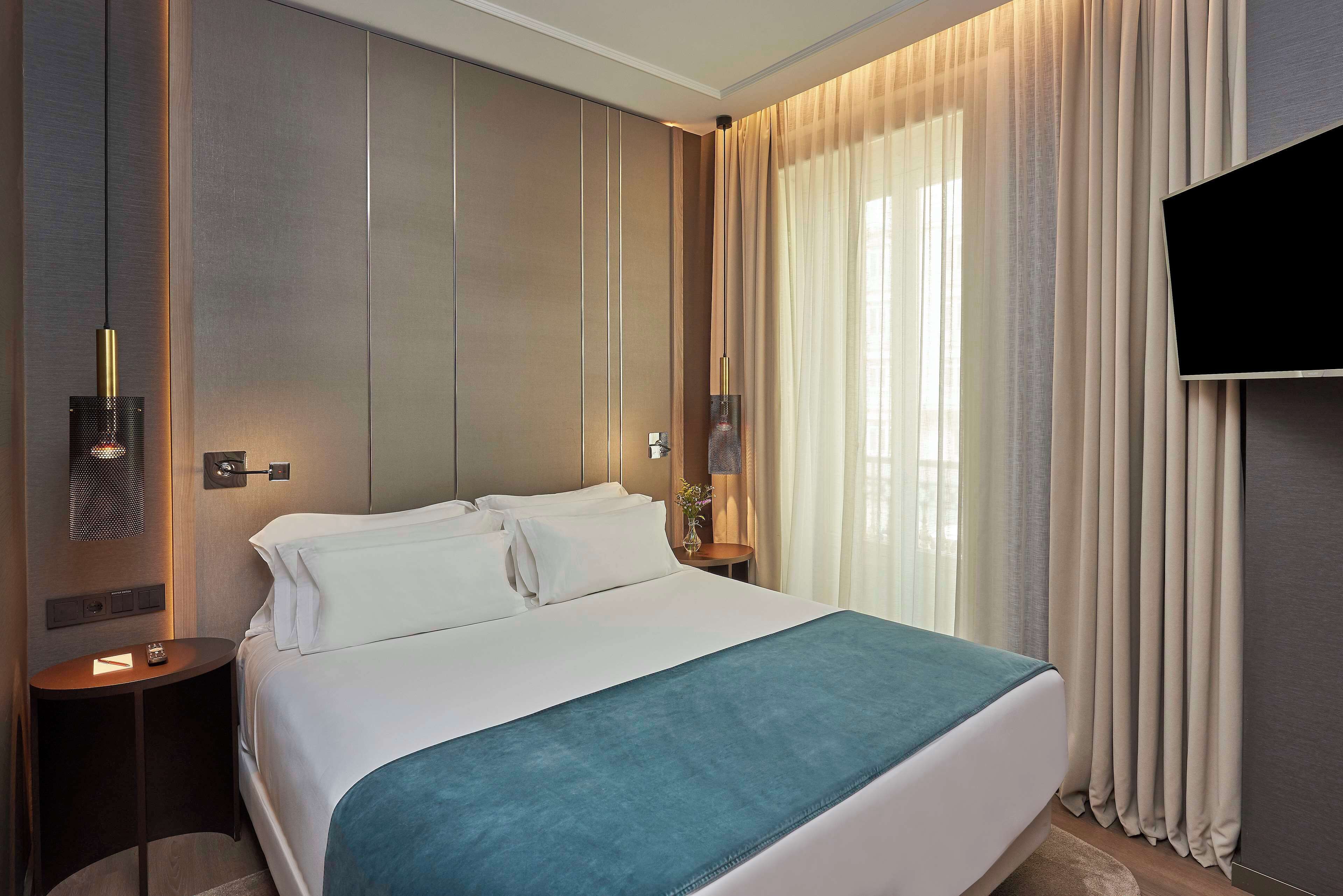 Quarto de hotel com cama, televisão presa na parede e mesa de cabeceira com abajur.