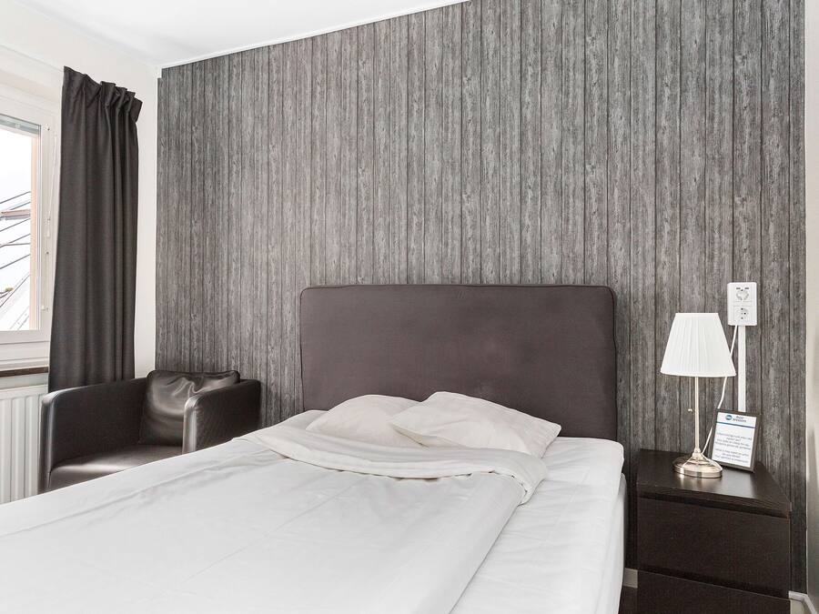 Cama bem arrumada com lençóis brancos, cabeceira escura, luminária de cabeceira e uma poltrona de couro.