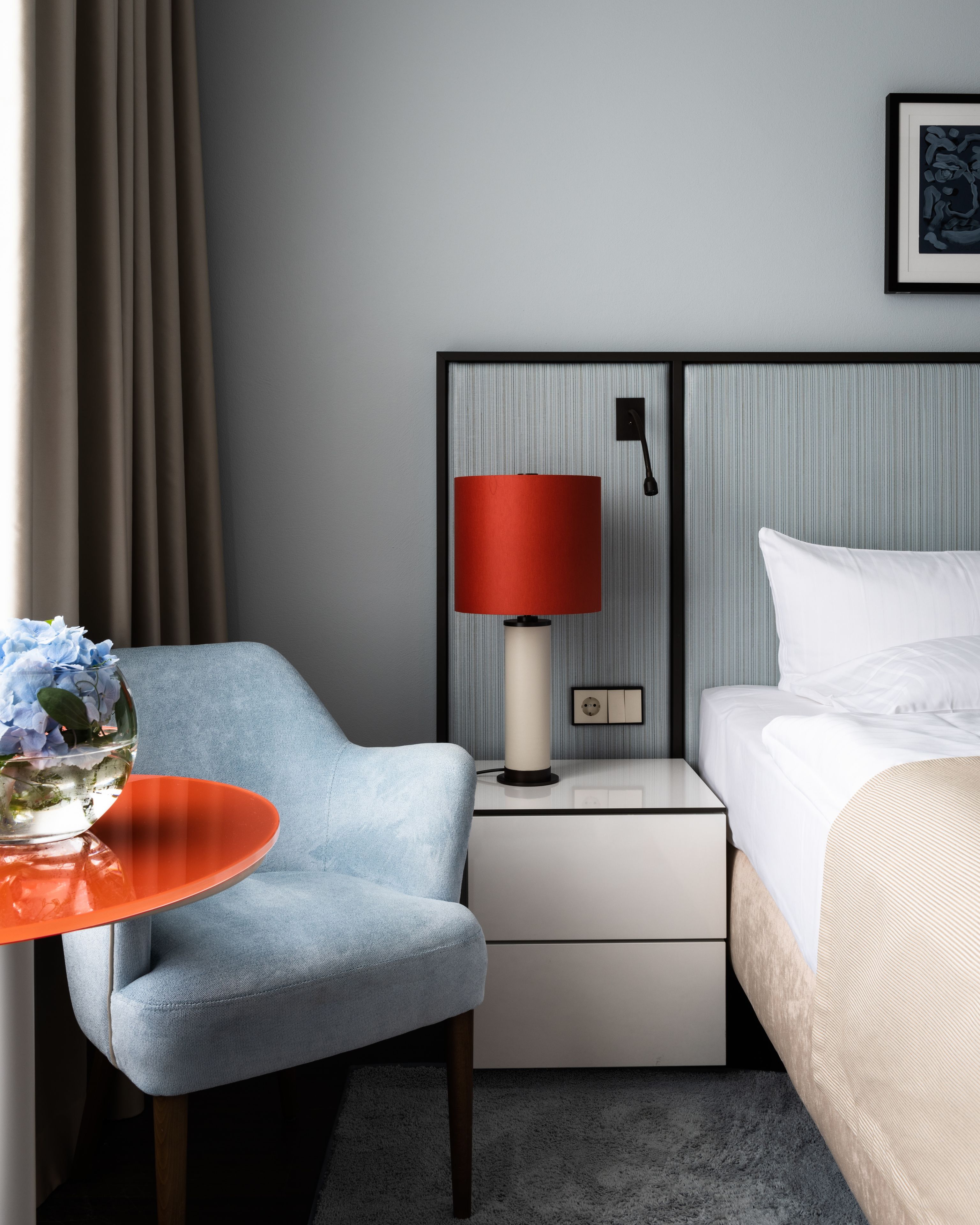 Quarto de hotel com cama, mesa de cabeceira com abajur, uma cadeira e uma pequena mesa com um vaso de flores.