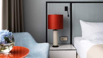 Quarto de hotel com cama, mesa de cabeceira com abajur, uma cadeira e uma pequena mesa com um vaso de flores.