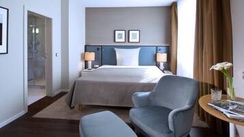 Quarto de hotel com cama, uma cadeira, mesa com uma revista e um vaso com flor.