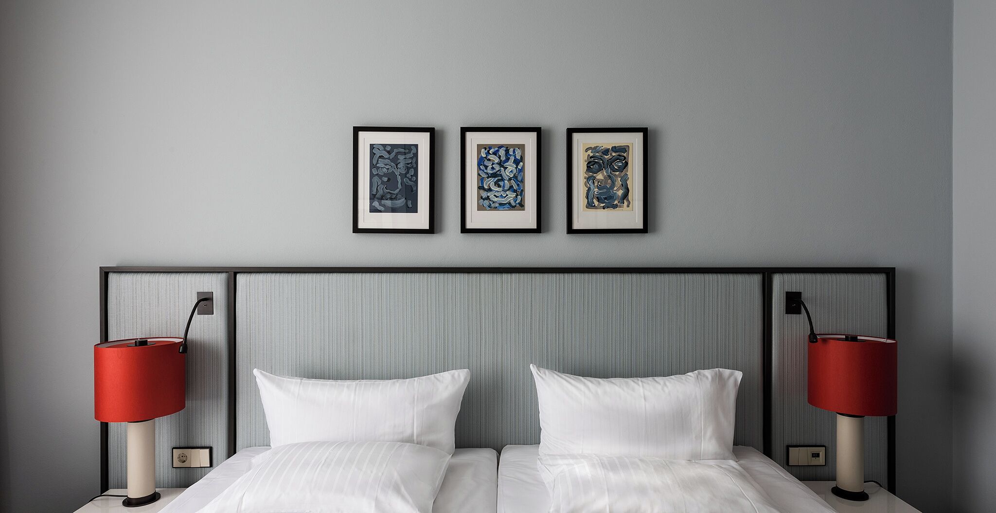 Quarto de hotel com uma cama, duas luminárias vermelhas e três obras de arte emolduradas na parede.