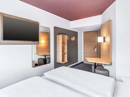 Quarto de hotel moderno com uma cama, televisão fixada na parede, guarda-roupa de madeira e um pequeno espaço de estar com mesa e cadeiras.