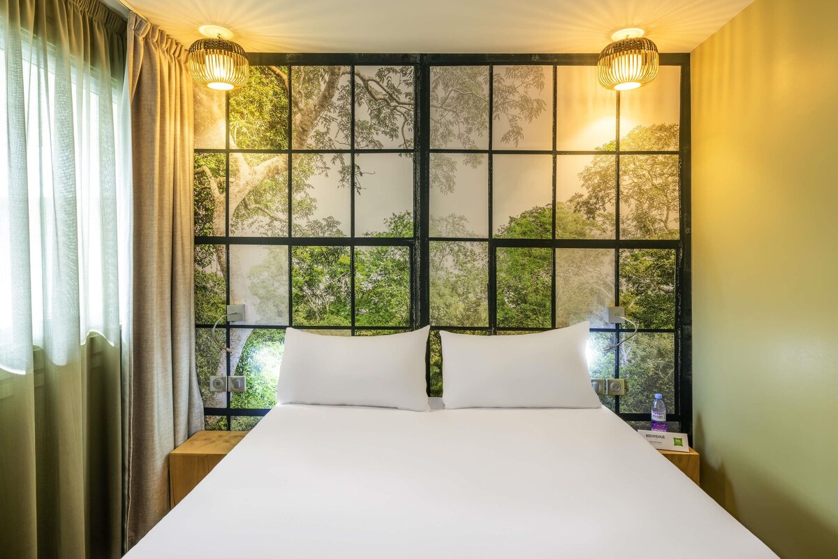 Quarto de hotel com uma cama grande, cortinas, uma janela com vista para árvores e duas luminárias fixadas na parede.