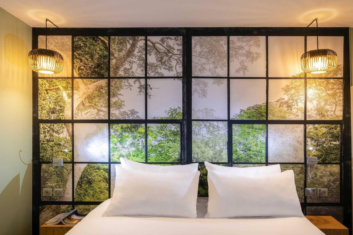Cama com roupa de cama branca, mesa de cabeceira com abajur e janela com vista para árvores.