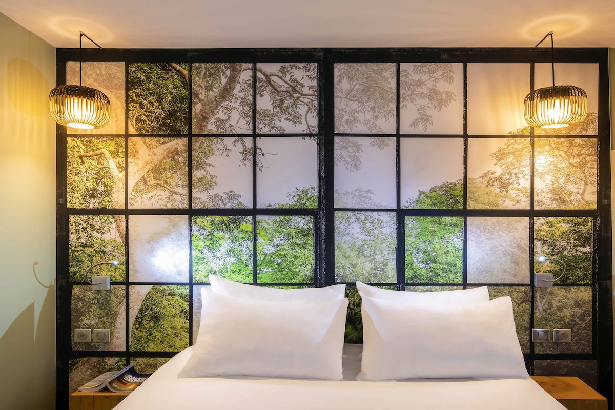 Uma cama com lençóis brancos, uma mesa de cabeceira e um abajur fixado na parede.