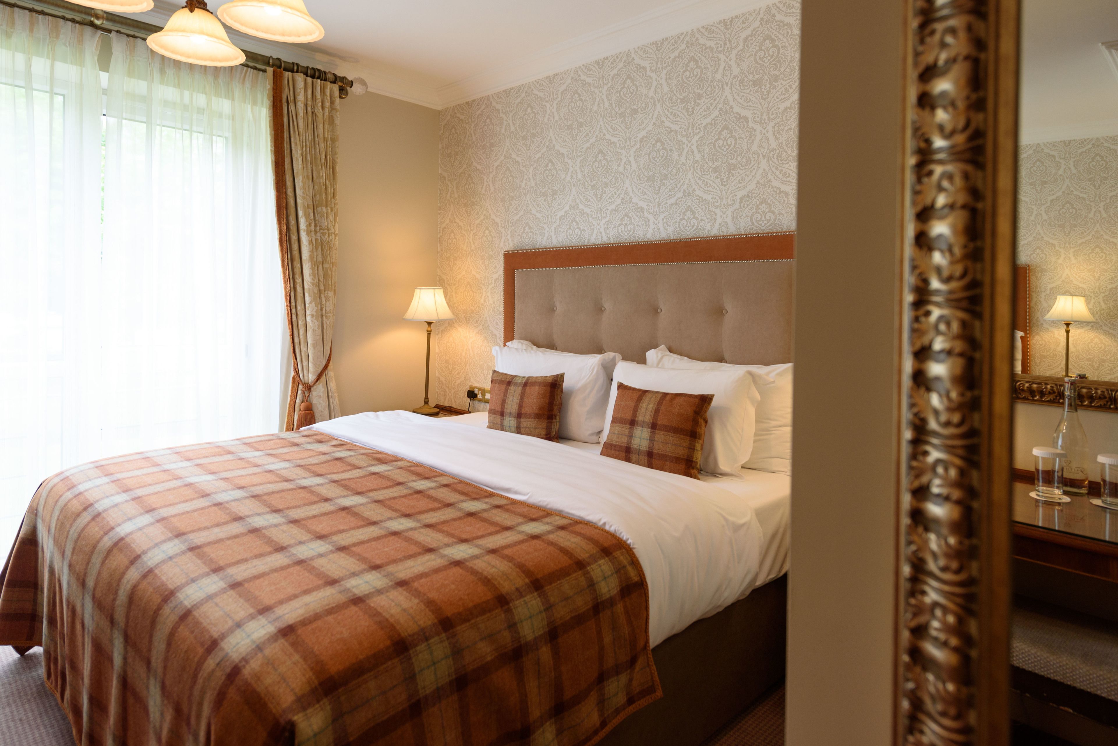 Quarto de hotel com uma cama grande, cabeceira, duas mesas de cabeceira com abajures, um espelho e uma janela com cortinas.