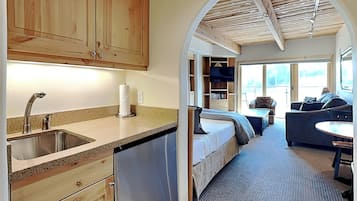 Cozinha com armários de madeira, pia de aço inoxidável e bancada. Há uma cama com cobertor azul e um sofá na sala de estar.