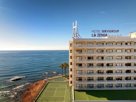 Um hotel com vários andares, varandas, quadra de tênis e vista para o mar.