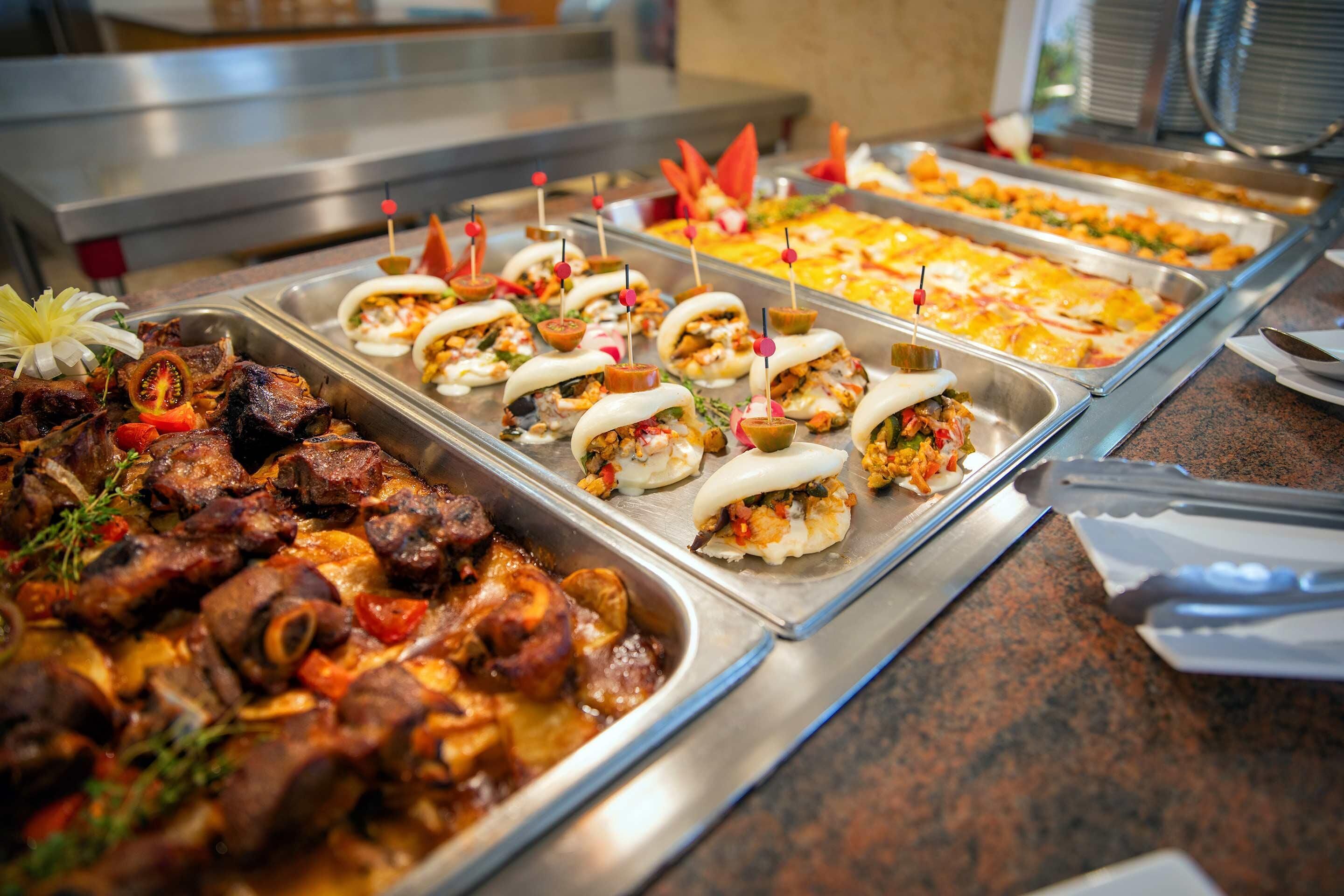 Buffet com diversos pratos, incluindo carne, legumes e mini pães.