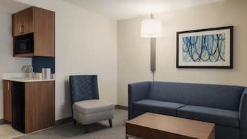 Quarto de hotel moderno com cozinha compacta, um sofá cinzento, uma poltrona azul, uma mesa de centro de madeira e uma pintura abstrata emoldurada na parede.