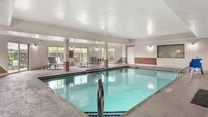 Piscina interior com água cristalina, rodeada por espreguiçadeiras e mesas, e grandes janelas com vista para o verde.