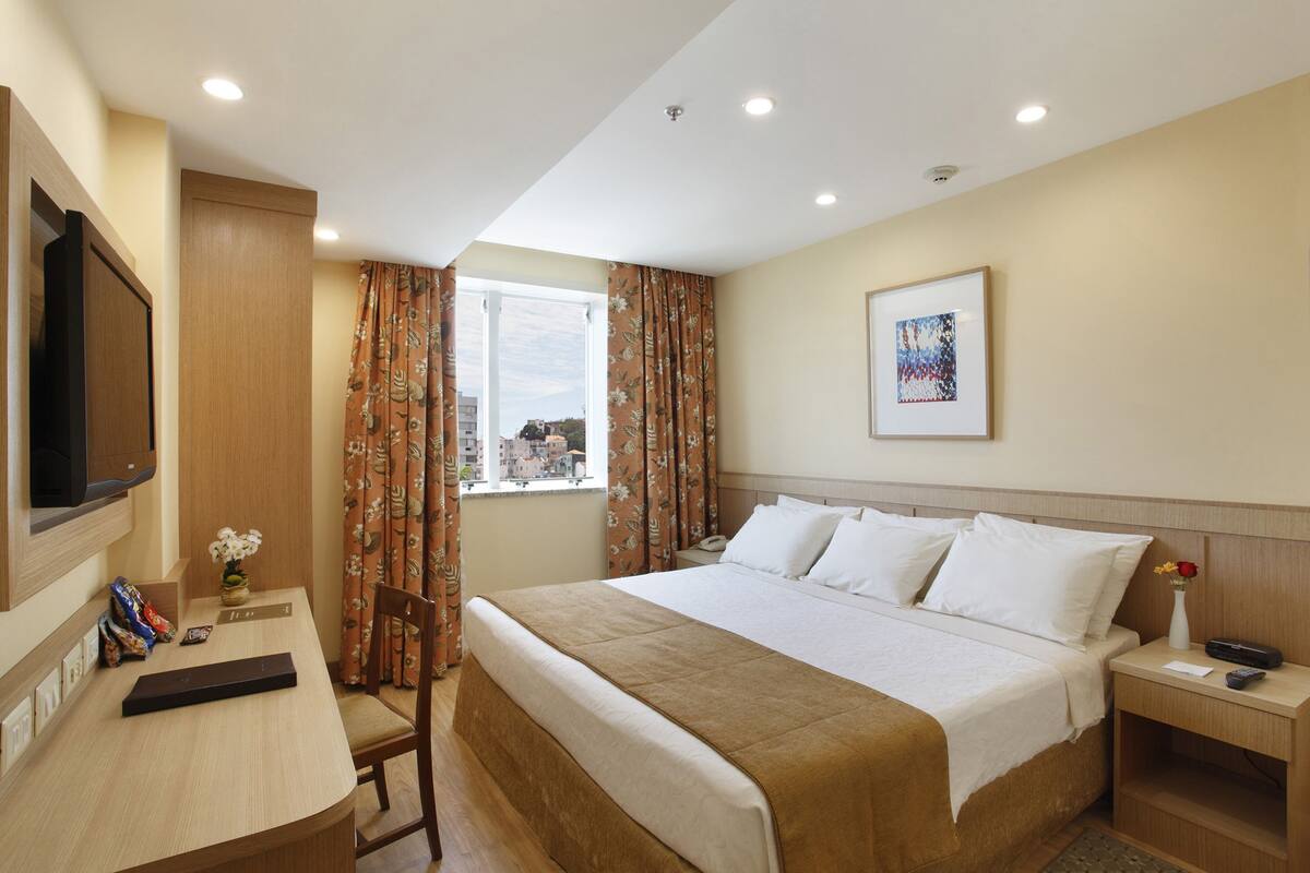 Quarto de hotel com cama, mesa de cabeceira, televisão e janela com cortinas.
