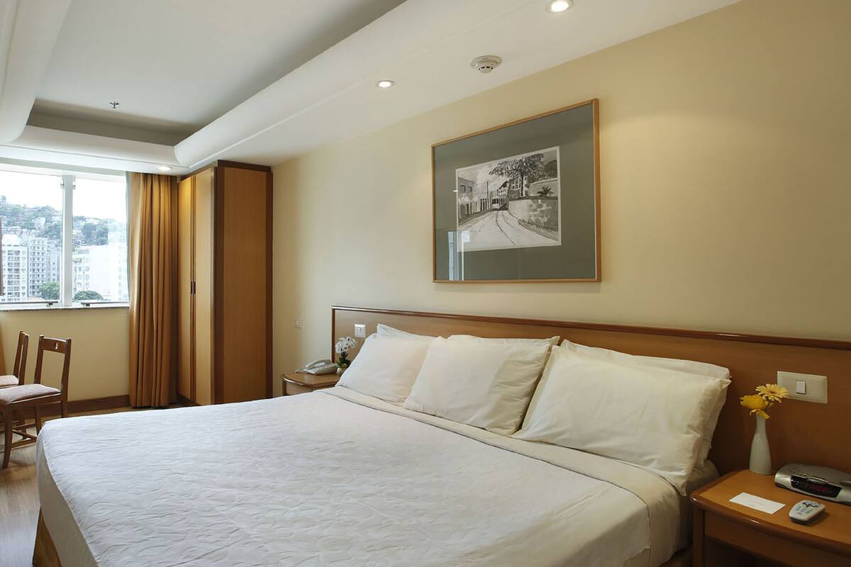 Quarto de hotel com uma cama grande, mesa de cabeceira com telefone e um vaso, uma cadeira e uma janela com cortinas.