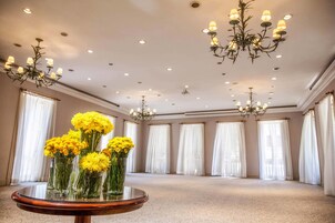 Um quarto espaçoso com um grande candelabro, várias janelas com cortinas e uma mesa redonda com vasos de flores amarelas.