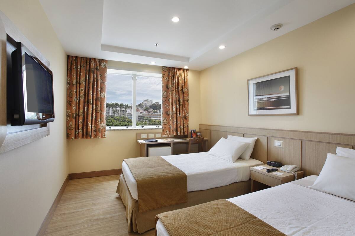 Quarto de hotel com duas camas, uma escrivaninha, televisão e janela com cortinas.