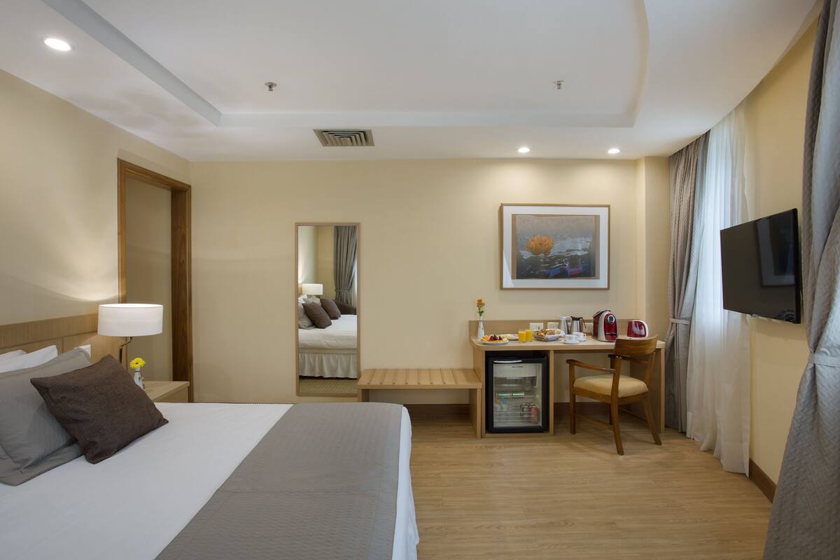 Quarto de hotel com cama, mesa de cabeceira, luminária, espelho, escrivaninha com chaleira, cadeira e televisão fixada na parede.
