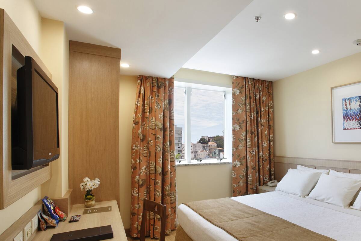 Quarto de hotel com uma cama, escrivaninha com cadeira, televisão e janela com cortinas.