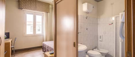 Quarto com cama, escrivaninha e cadeira. Há uma janela com cortinas e vista para edifícios.