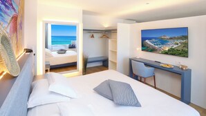 Quarto de hotel moderno com cama, escrivaninha, cadeira, televisão e pintura de uma paisagem costeira.
