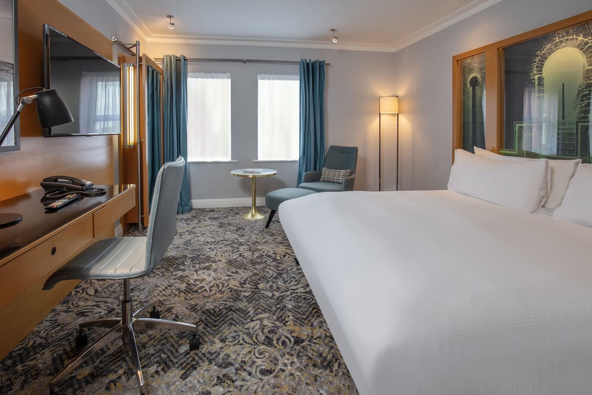Quarto de hotel com uma cama grande, uma escrivaninha com telefone, uma cadeira, uma mesinha redonda e um espelho amplo.