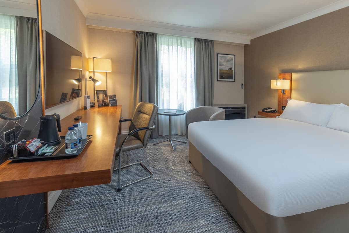 Quarto de hotel com uma cama grande, uma escrivaninha com cafeteira, uma cadeira, uma pequena mesa e um abajur.
