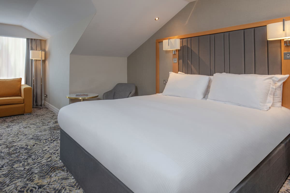 Quarto de hotel com uma cama grande, uma cadeira, uma mesa pequena e um abajur.