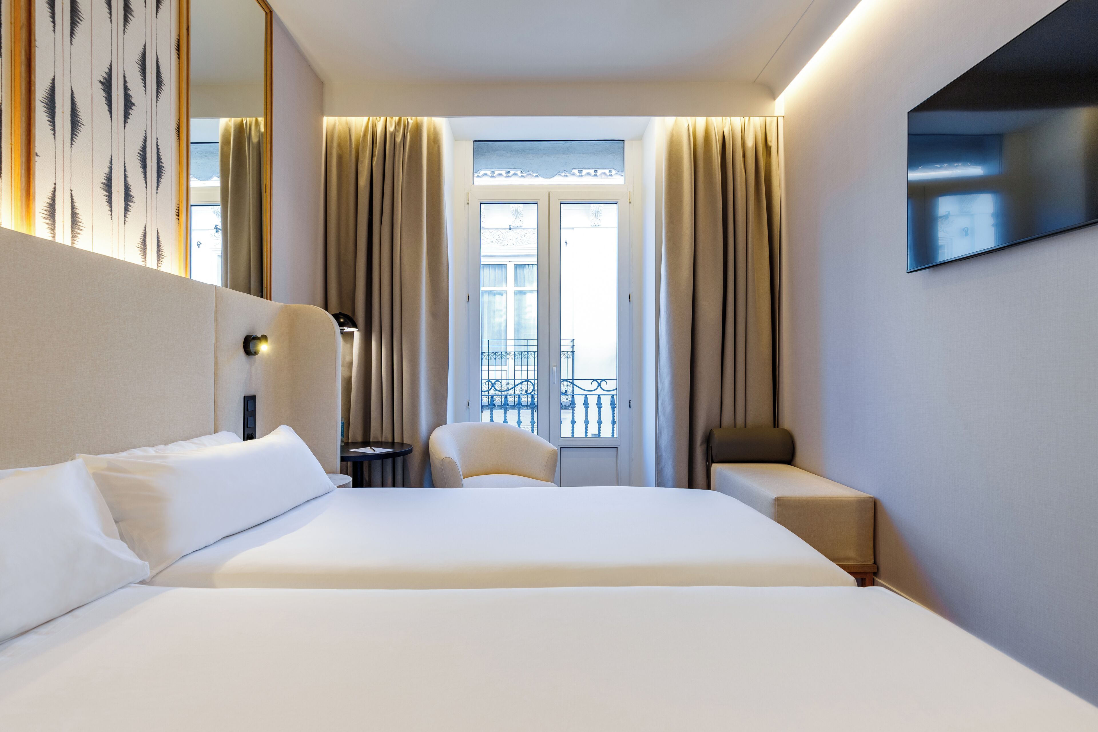 Quarto de hotel com uma cama grande, uma cadeira, uma mesinha e uma televisão fixada na parede.