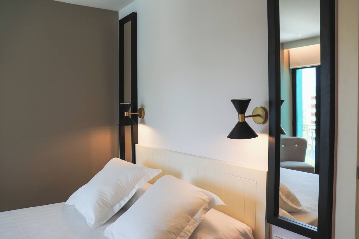 Quarto de hotel com cama, almofadas brancas, luminária fixada na parede e um espelho grande que reflete o quarto.