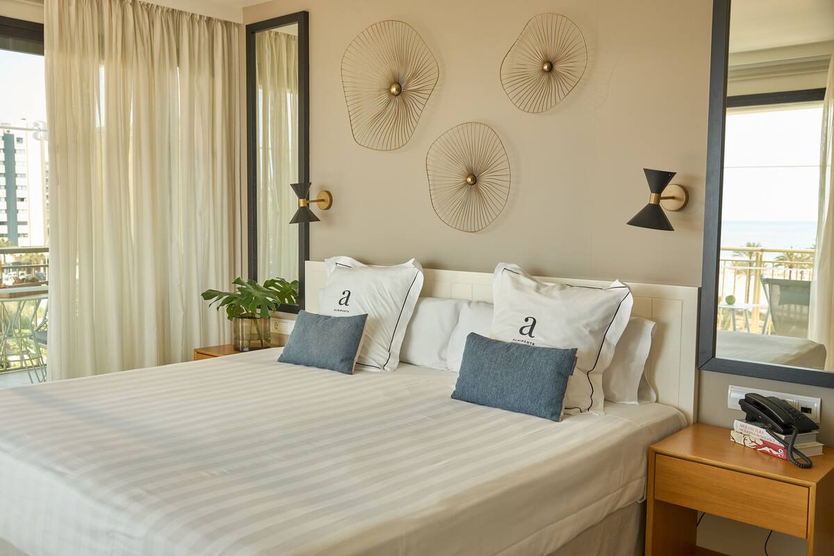 Quarto de hotel com uma cama grande, duas mesas de cabeceira, um criado-mudo com telefone e arte decorativa na parede.