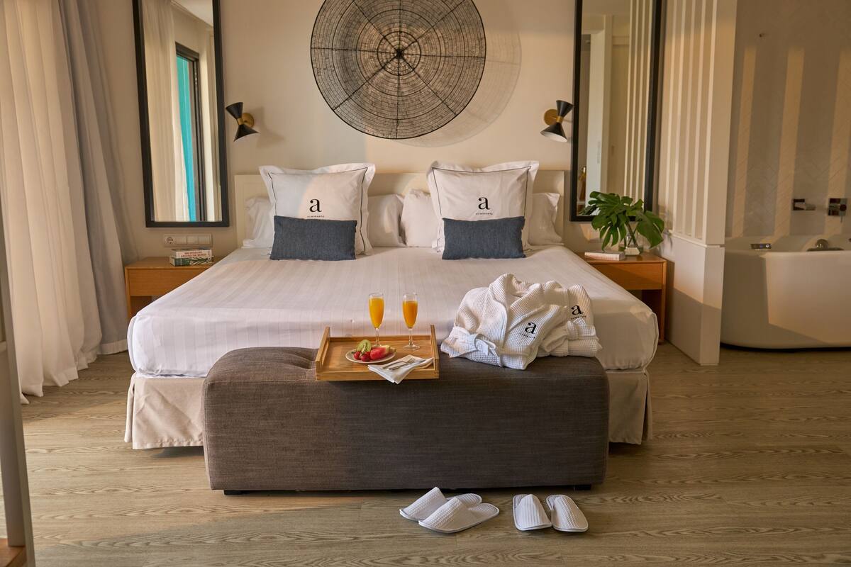 Cama bem arrumada com almofadas etiquetadas 'a', bandeja com fruta e suco, chinelos e roupão na cama.