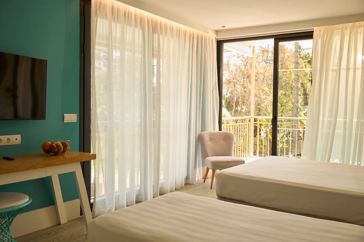 Quarto de hotel com duas camas, uma escrivaninha com televisão e varanda com uma cadeira.