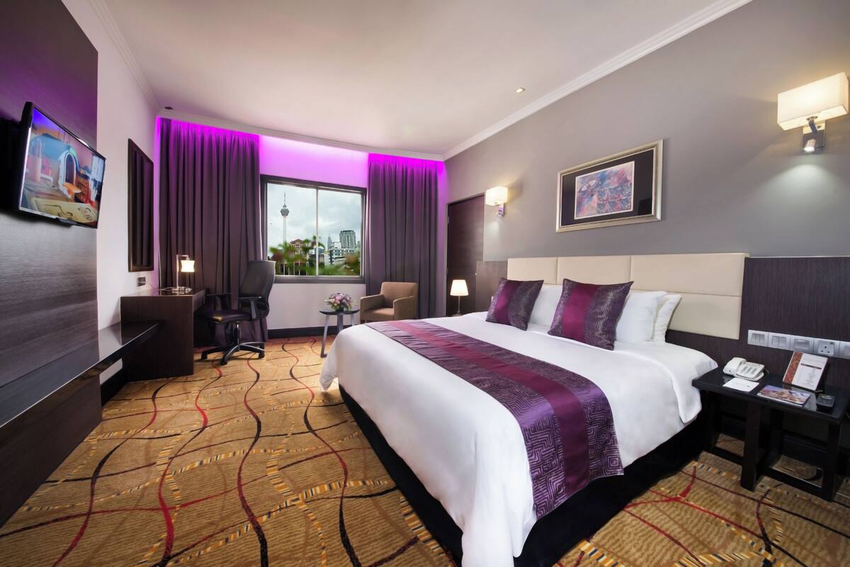 Quarto de hotel com uma cama grande, uma escrivaninha com telefone, uma cadeira, uma mesinha com abajur e uma TV fixada na parede.