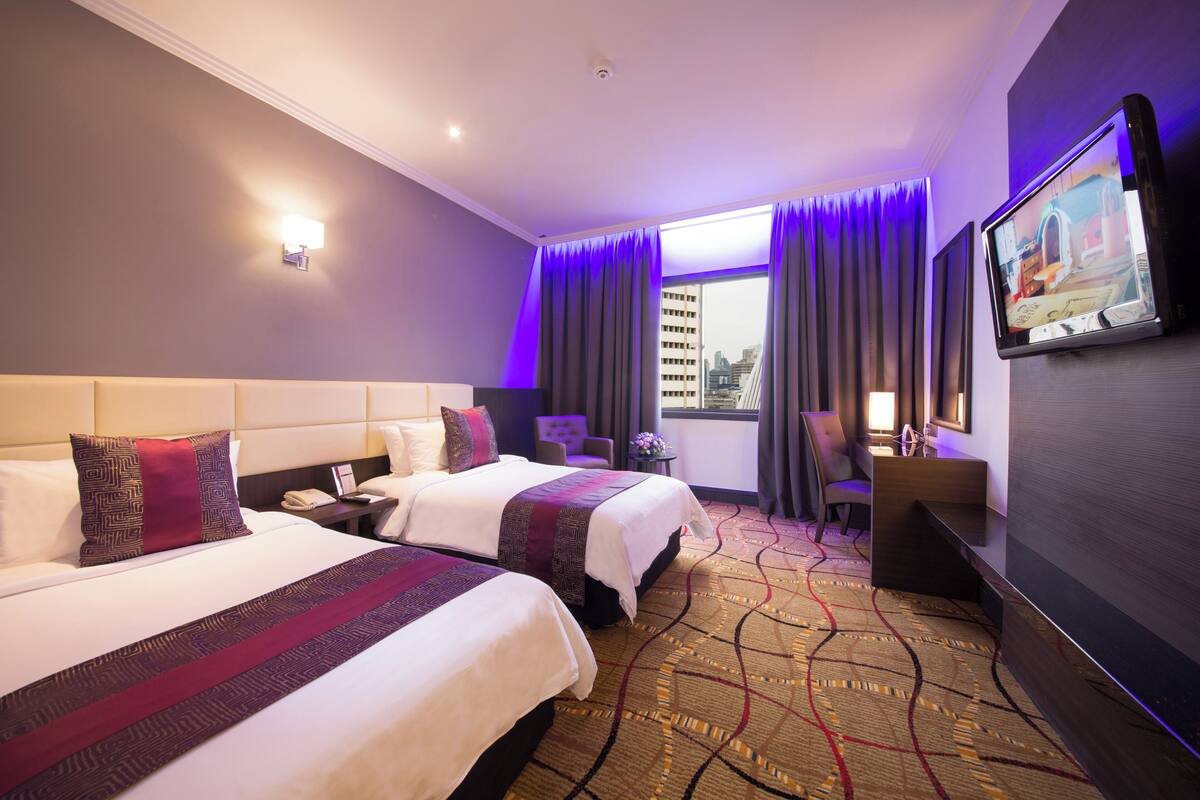 Quarto de hotel com duas camas, uma escrivaninha, uma cadeira, uma televisão e uma janela com cortinas.