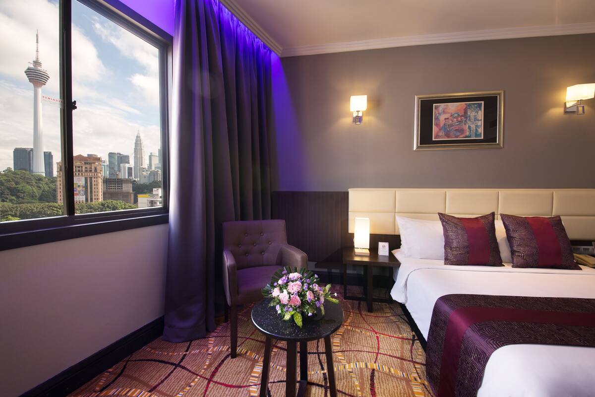 Quarto de hotel com uma cama grande, uma cadeira, uma mesa pequena com flores, um abajur e vista para a cidade.