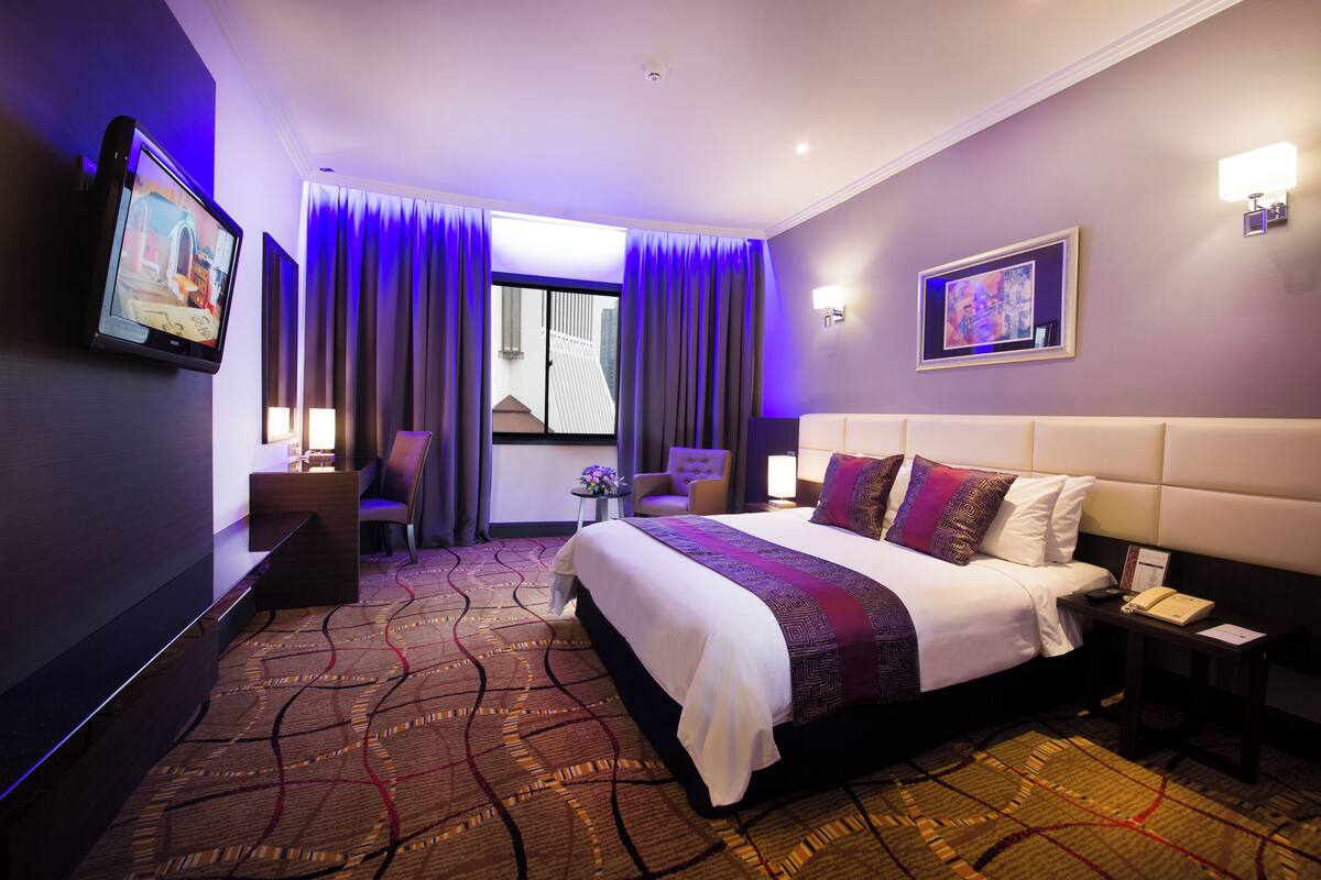 Quarto de hotel com uma cama grande, televisão, escrivaninha com cadeira, mesa pequena com luminária e janela com cortinas.