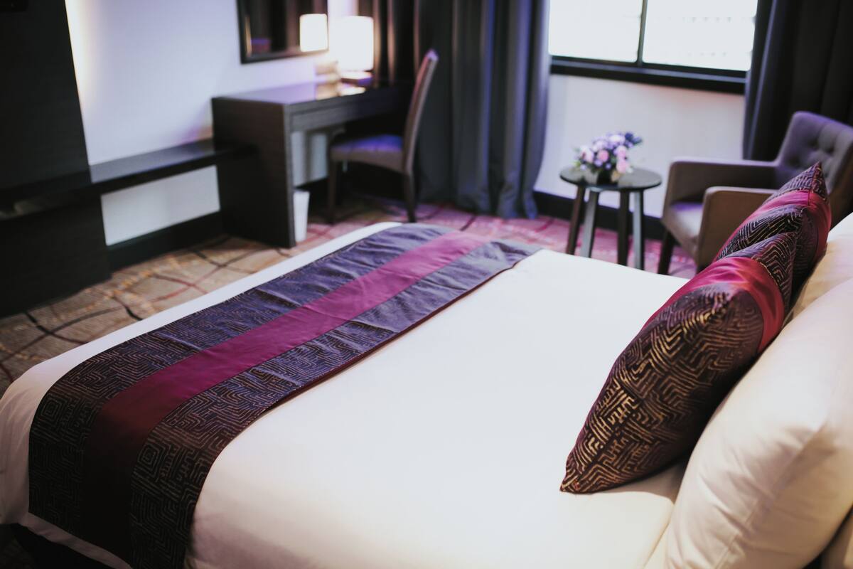 Quarto de hotel com cama, escrivaninha, cadeira e pequena mesa com flores.