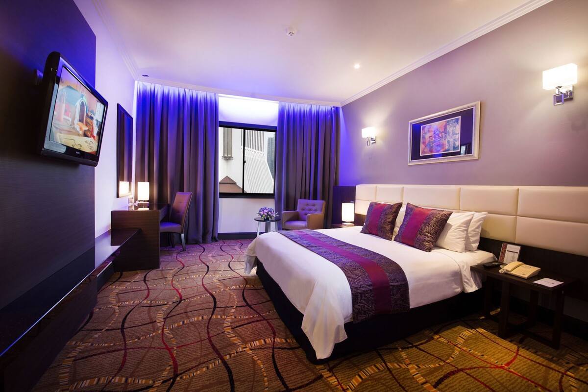 Quarto de hotel com uma cama grande, uma escrivaninha, duas cadeiras, uma televisão e uma janela com cortinas.